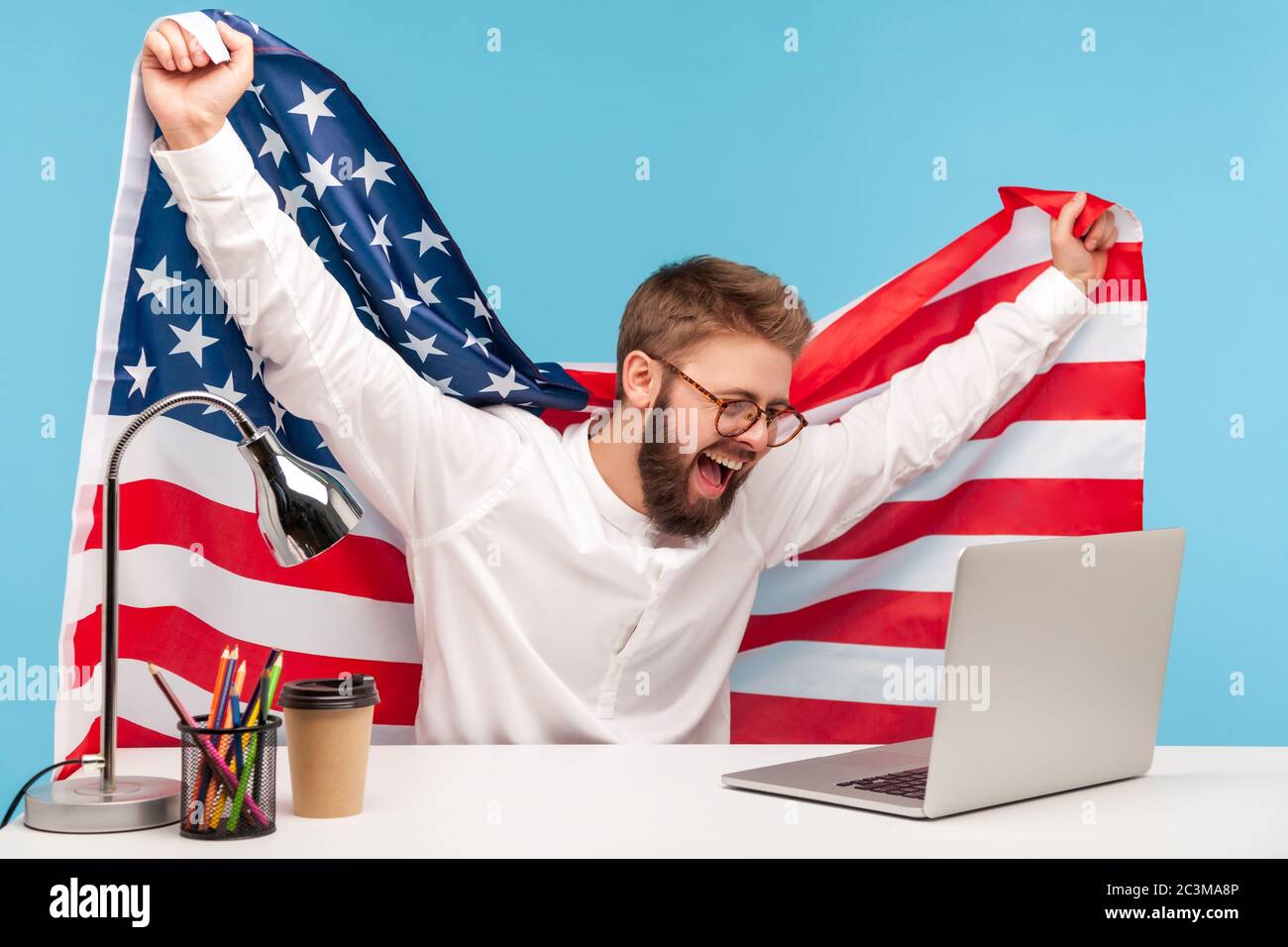 happy businessman raising American flag, watching live patriotic program on laptop at workplace ...