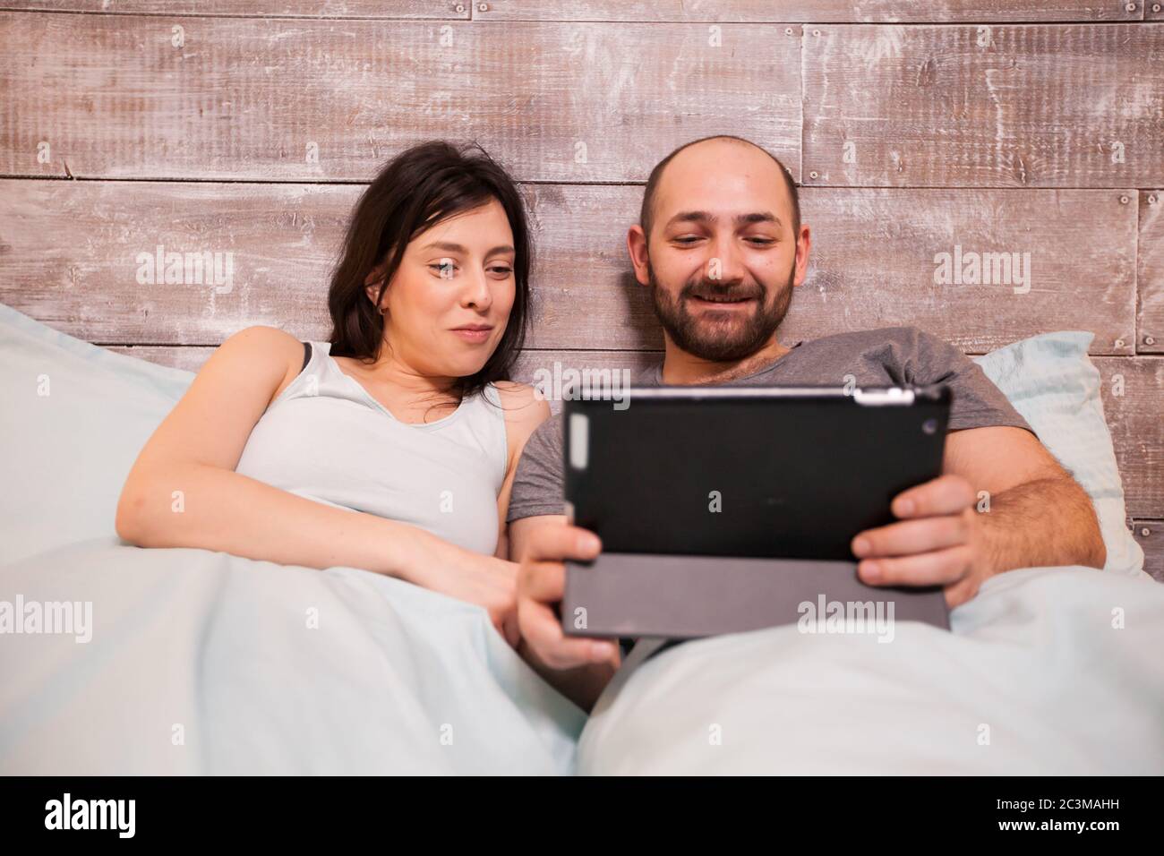 Beautiful young husband and wife wearing pajamas laughing while using ... Beautiful young husband and wife wearing pajamas laughing while using ...