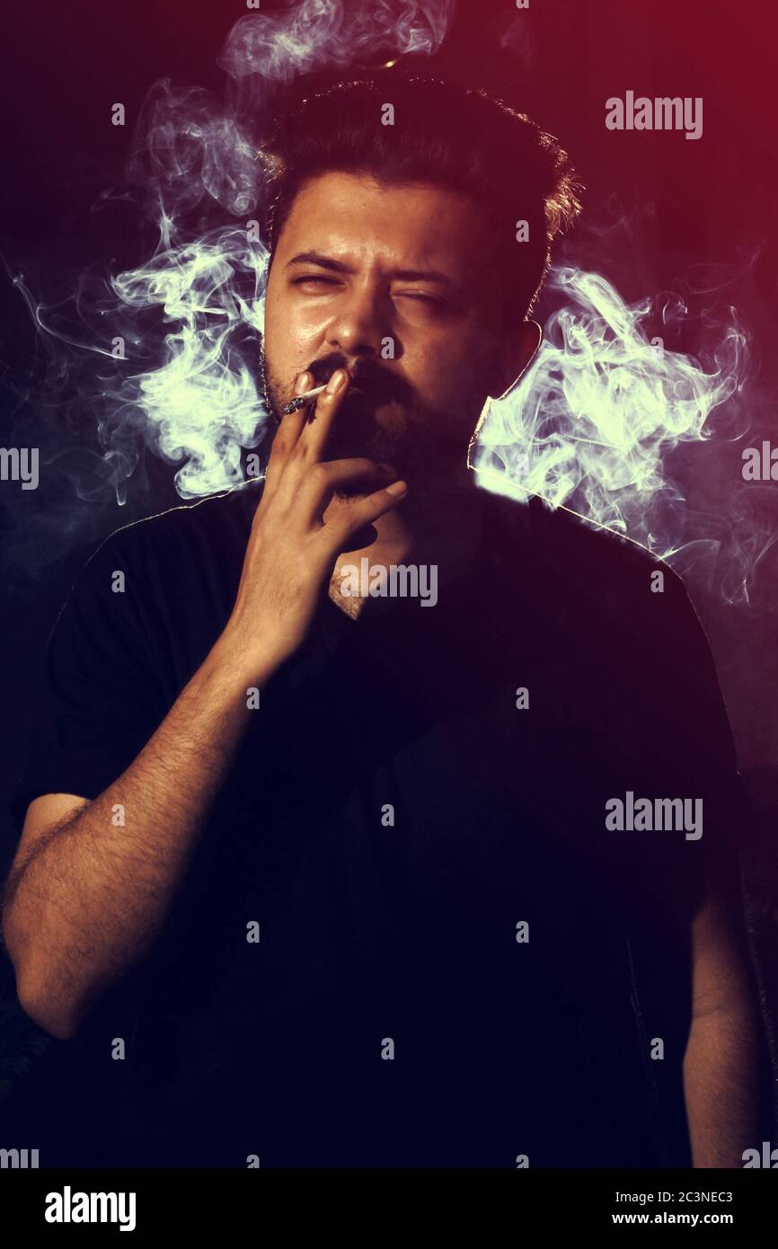 Young man smoking cigarette on dark background looking into camera ...