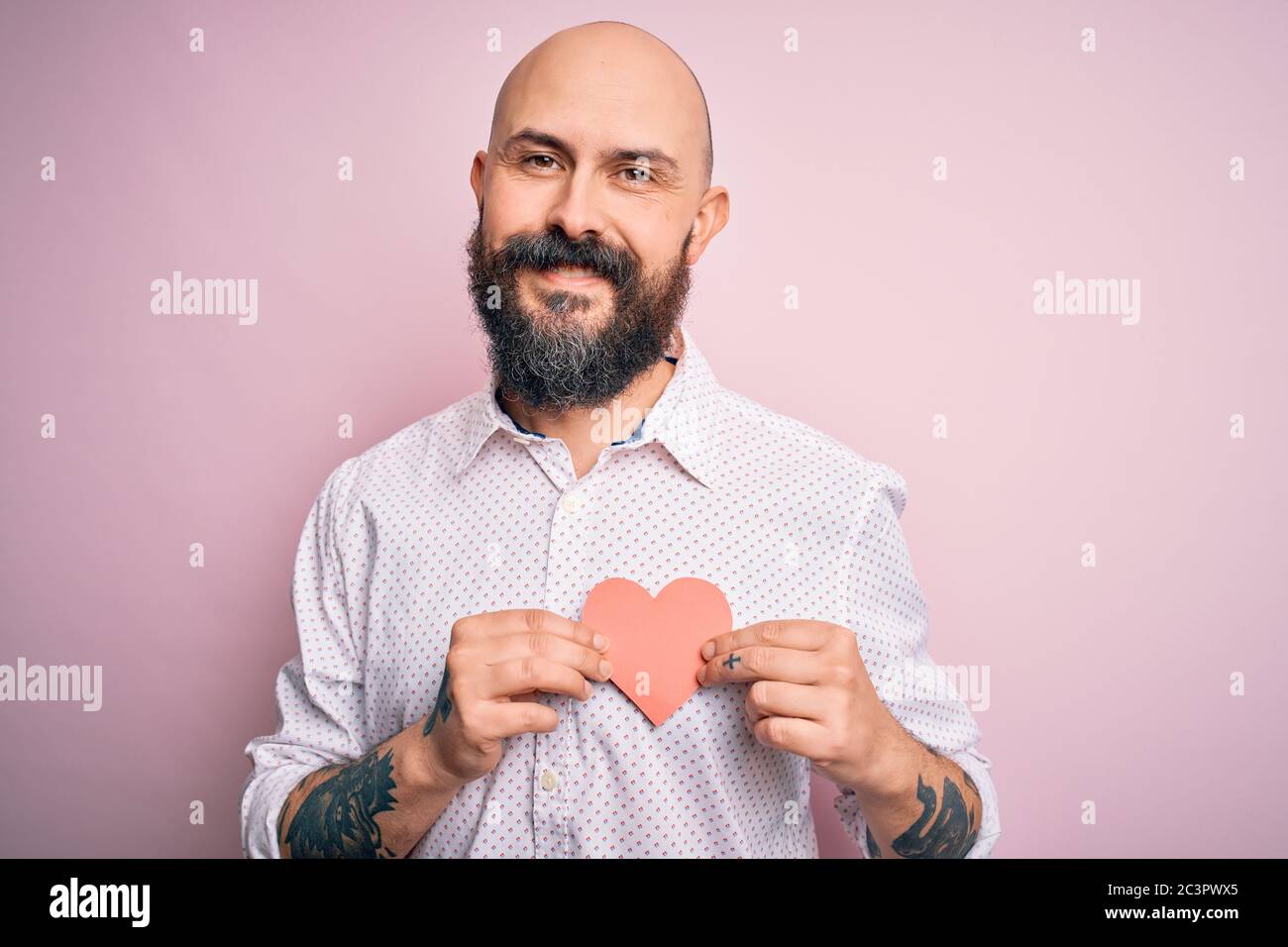 Handsome romantic bald man with beard holding red heart paper over pink ...