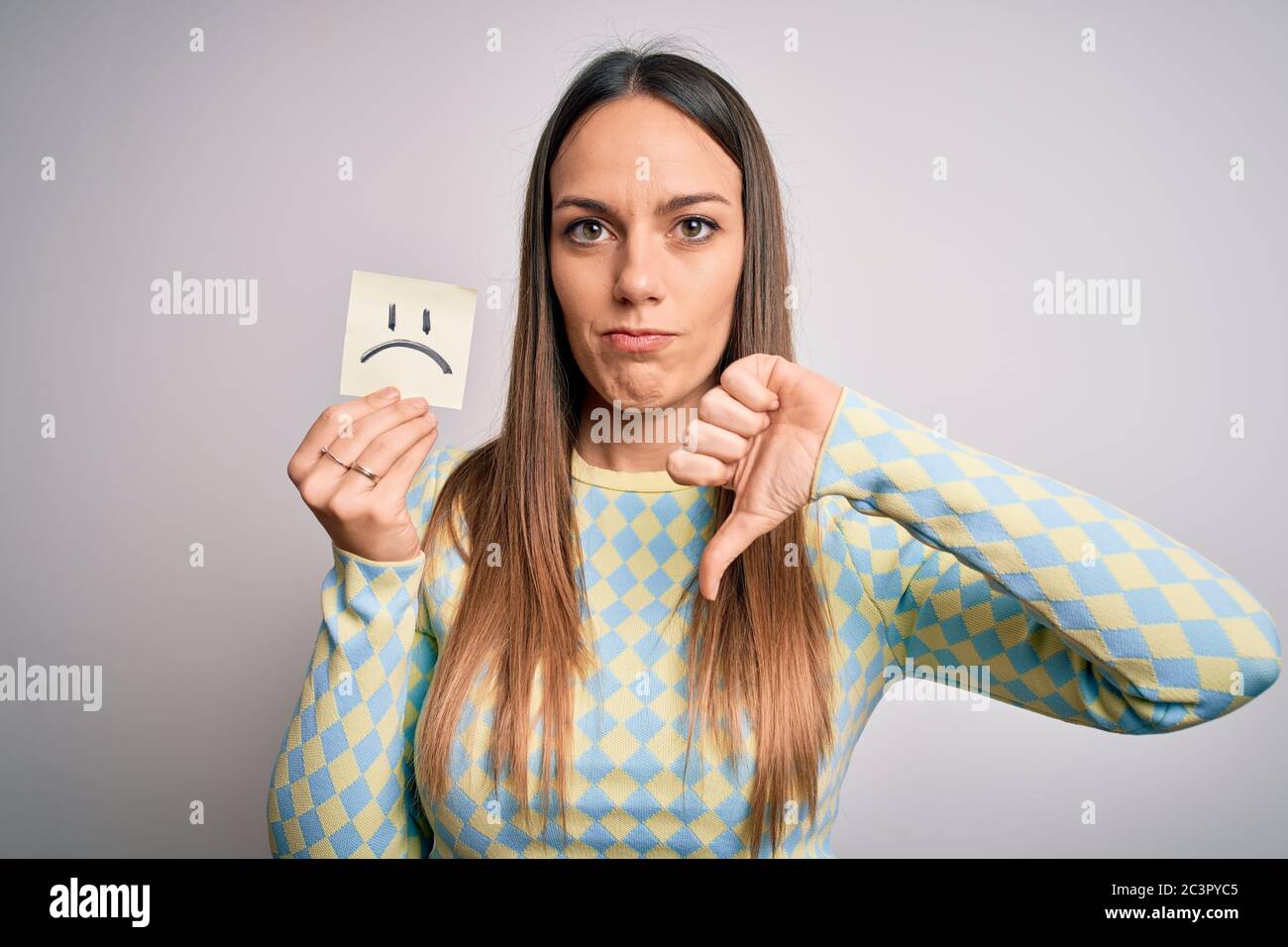 Young blonde woman holding sticky paper note with sad face emoticon ...
