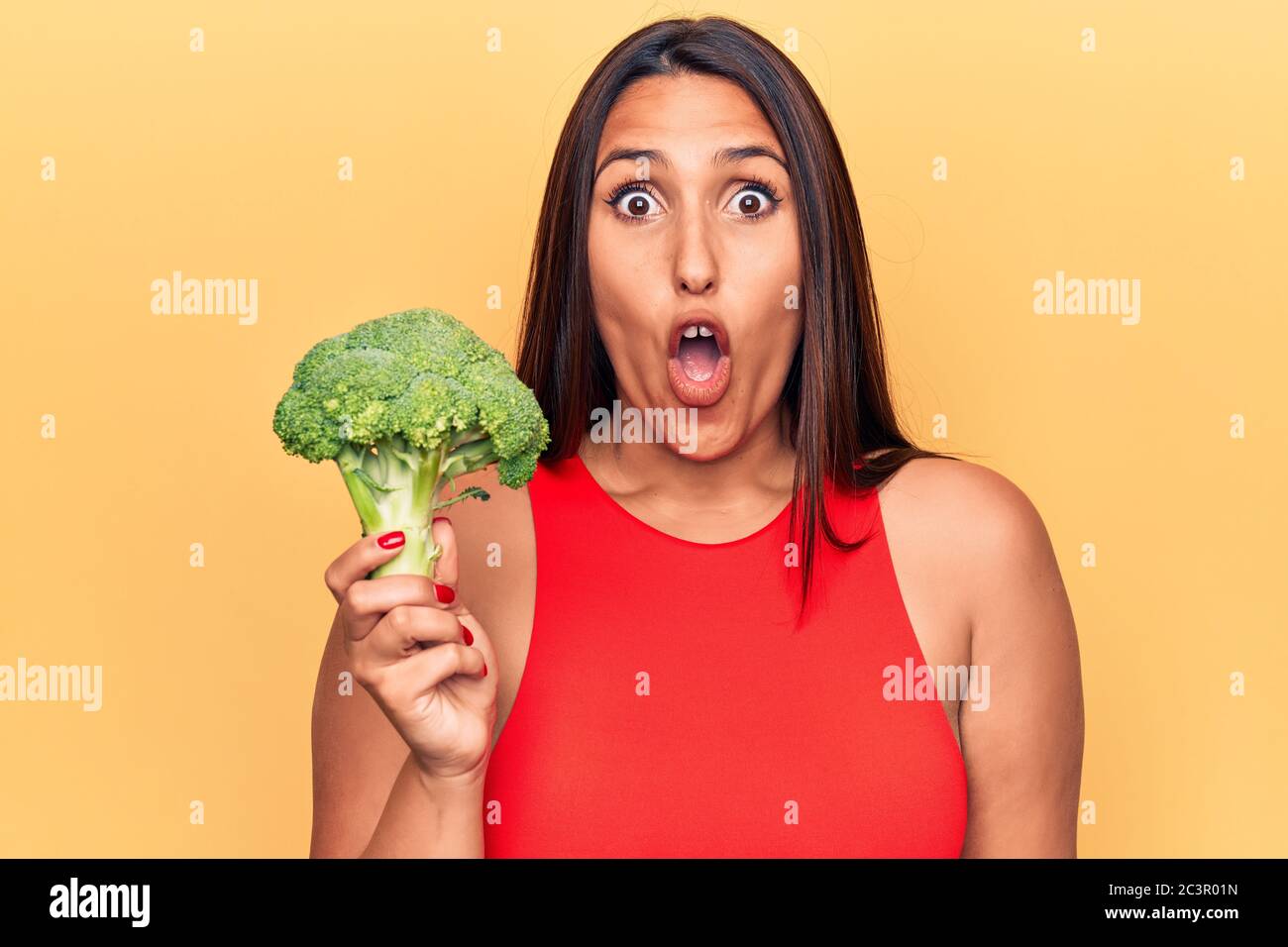 Young beautiful brunette woman holding broccoli scared and amazed with ...