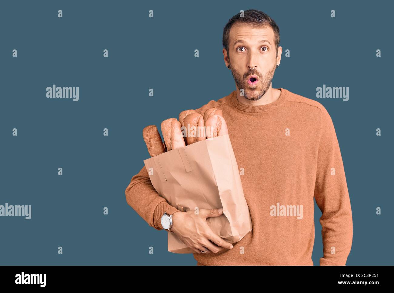 Young handsome man holding paper bag with bread scared and amazed with ...