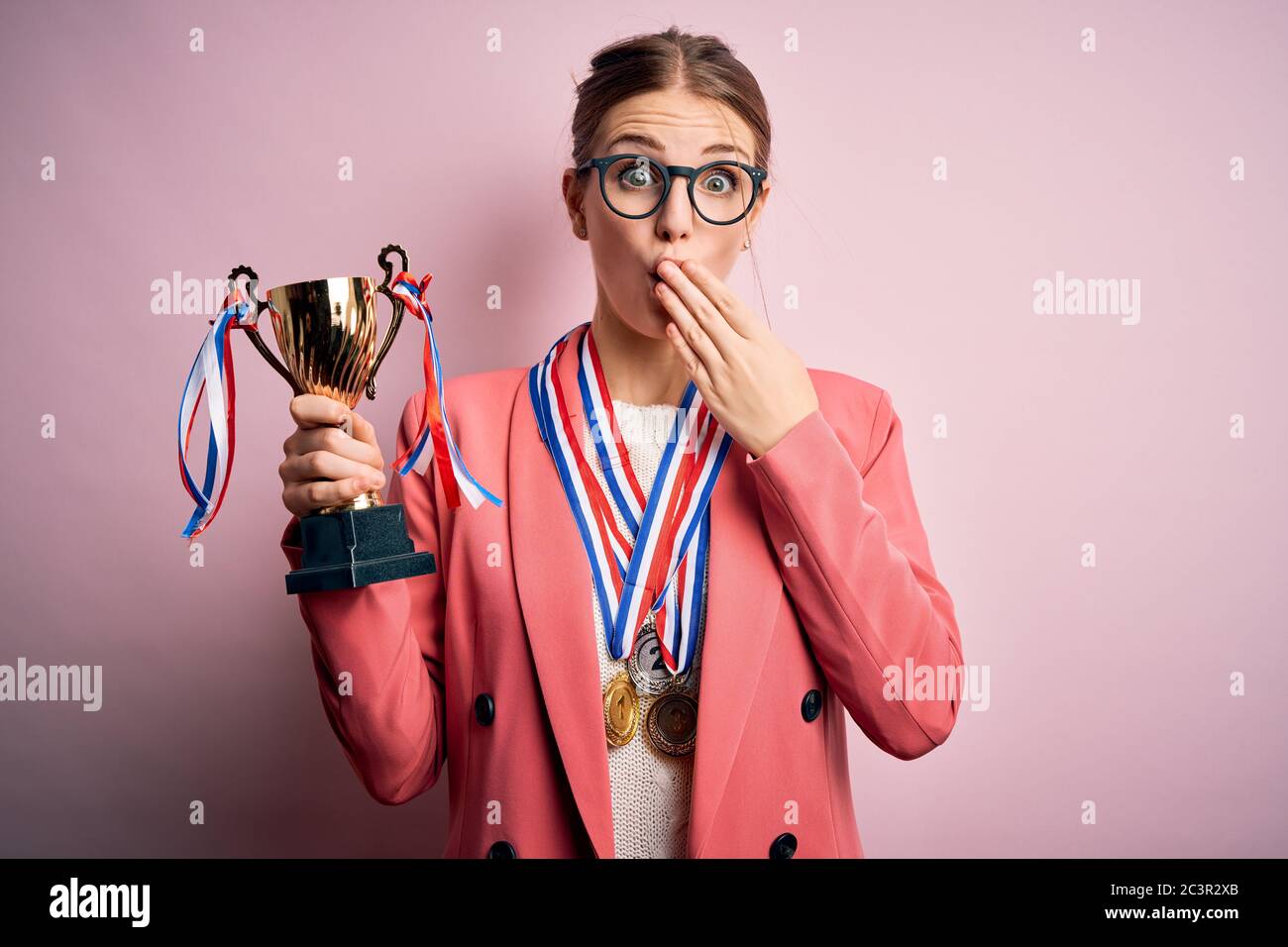 Young beautiful redhead woman holding trophy wearing medals over pink ...