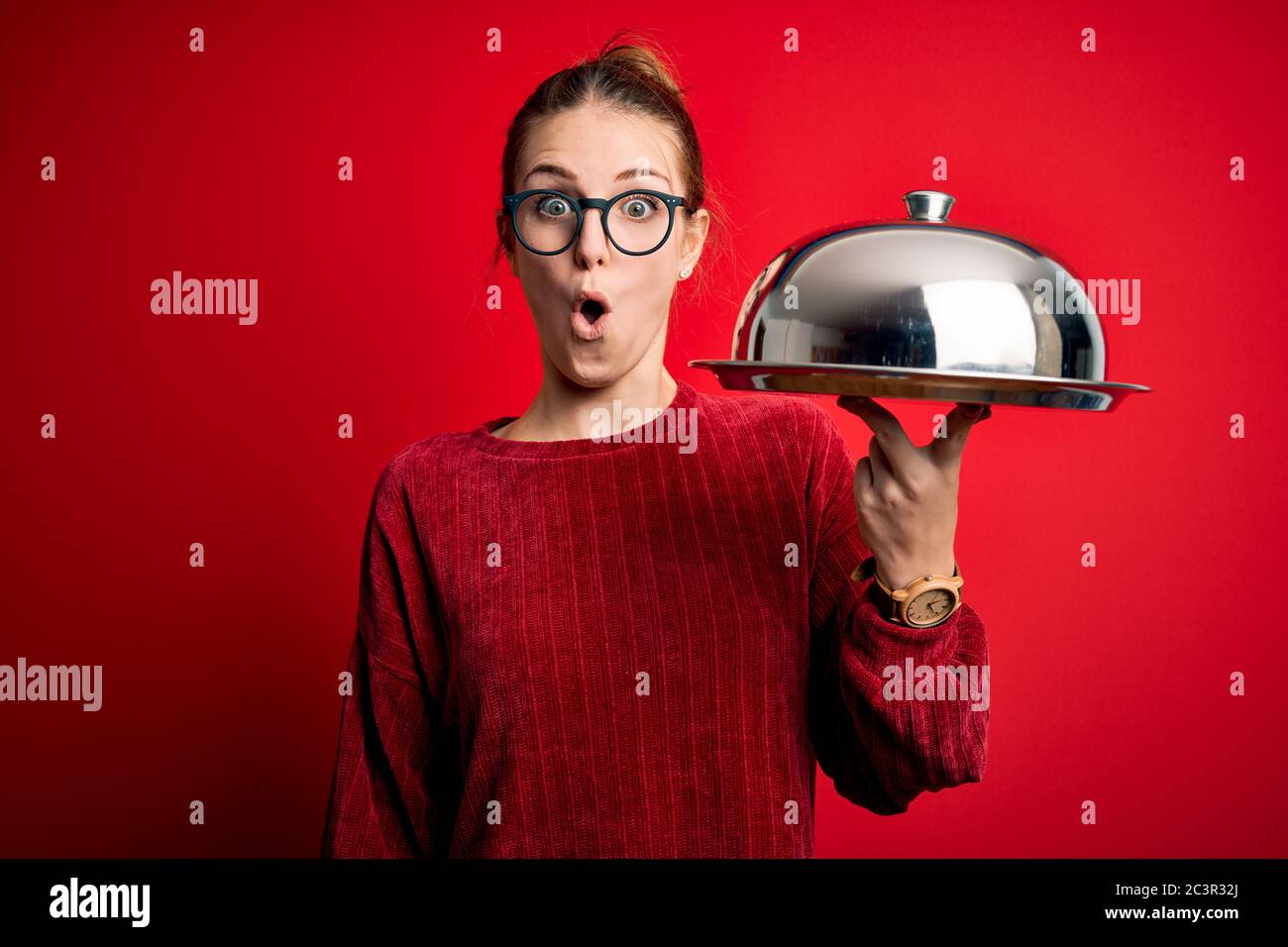 Young beautiful redhead woman holding waitress tray over isolated red ...