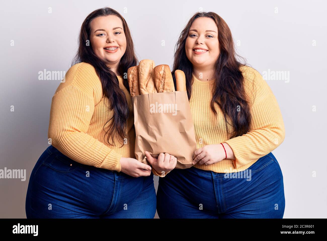 Young plus size twins holding paper bag with bread looking positive and ...