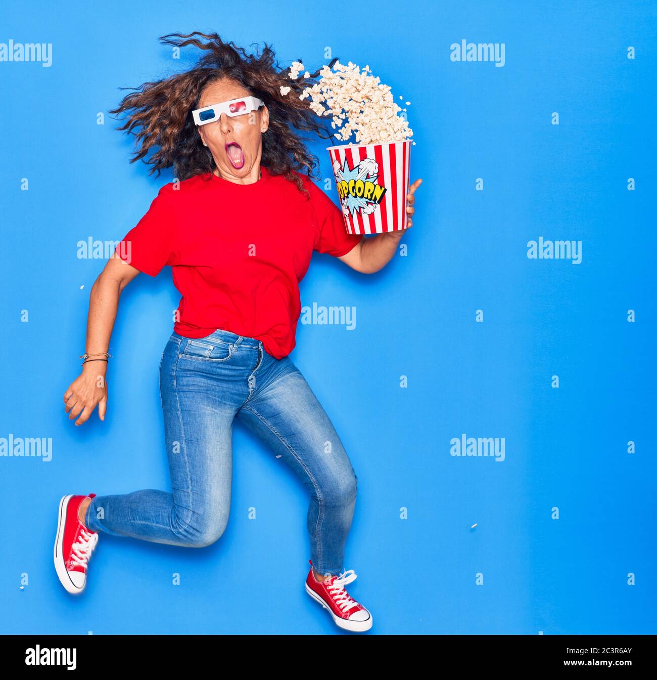 Middle age beautiful hispanic woman surprised with open mouth wearing 3d glasses. Holding bucket ...