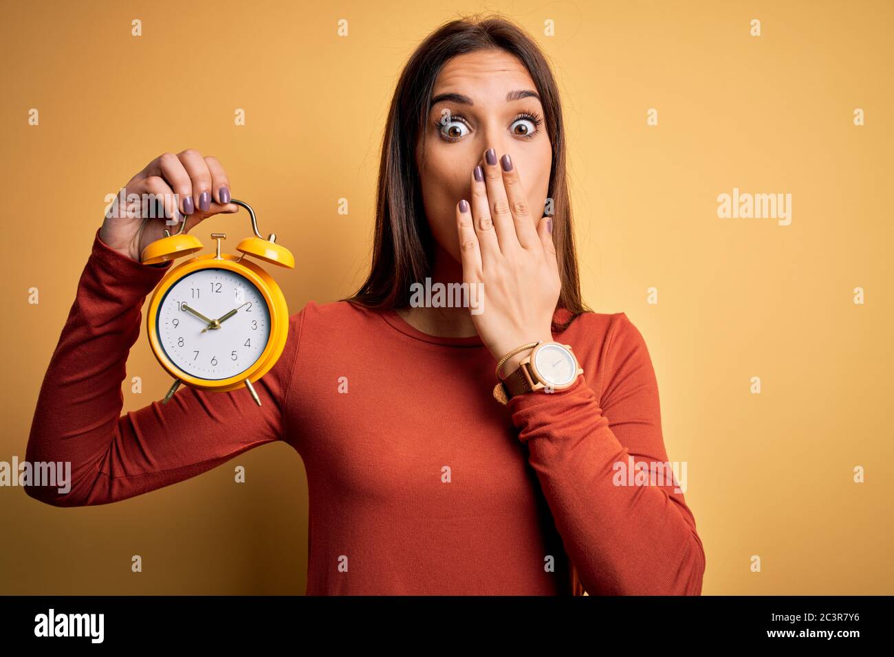 Young beautiful brunette woman holding alarm clock over isolated yellow ...