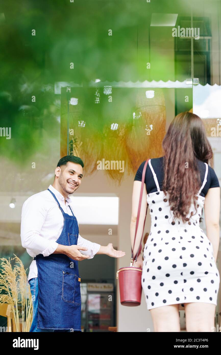 Positive young man in denim apron making welcoming gesture and inviting ...
