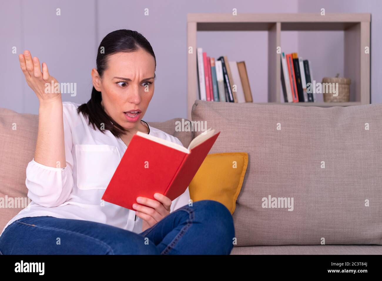Pretty young brunette woman confused reading a book sitting on the sofa ...