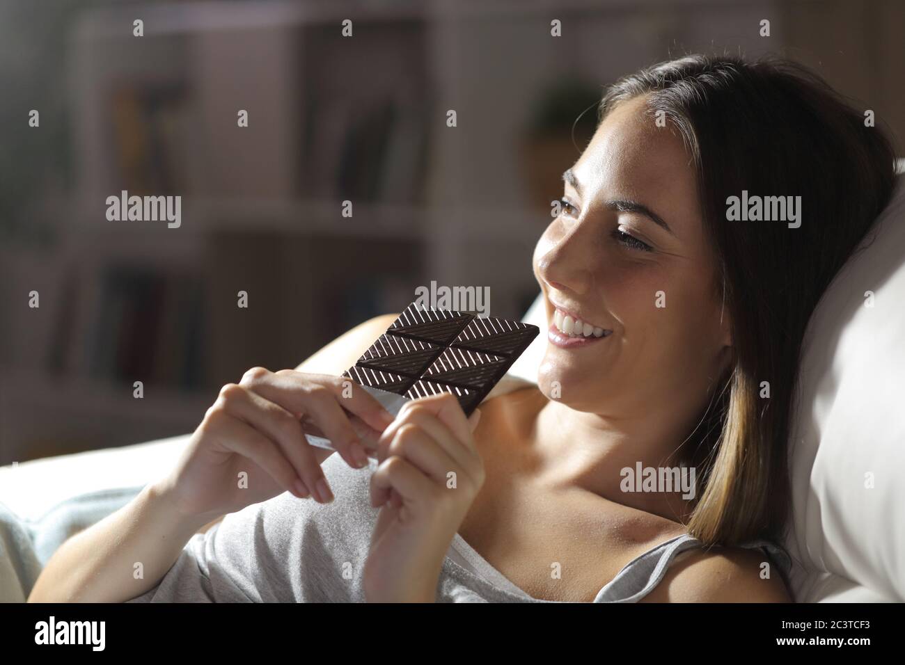 Happy girl eating chocolate bar sitting on a couch at home at night ...