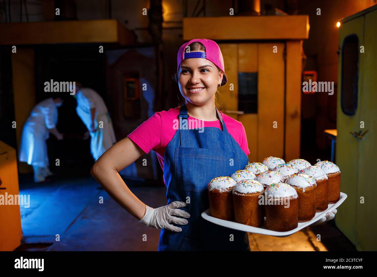 Girl baker holds a tray with hot pastries in the bakery. She is wearing ...