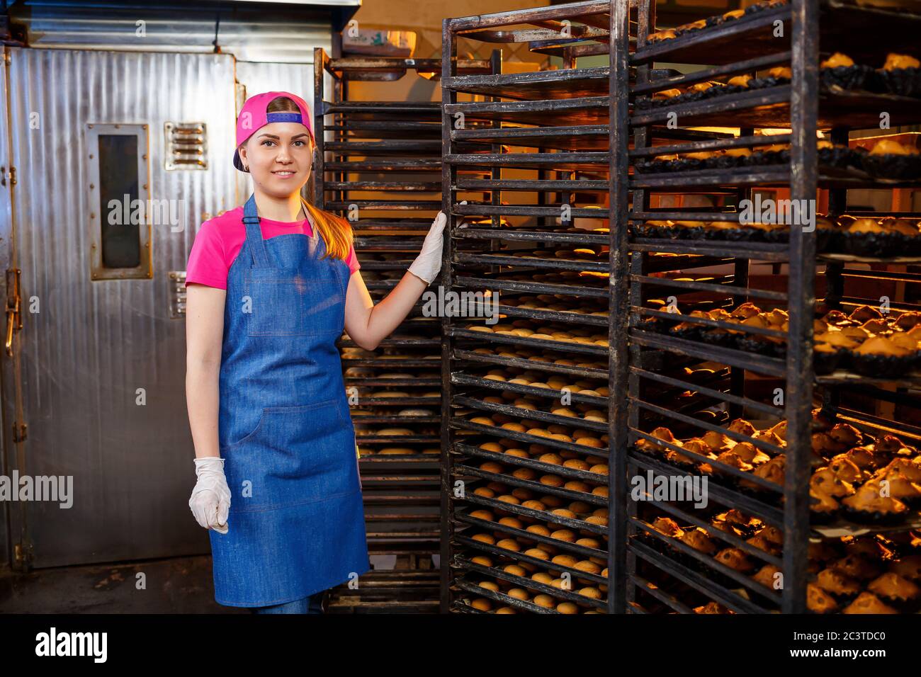 A professional baker girl is standing near shelving with trays of fresh ...