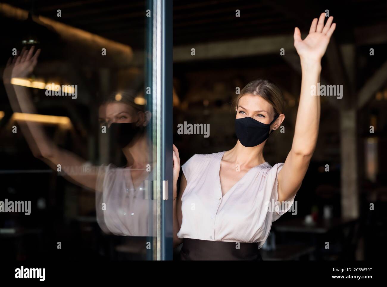 Portrait of waitress with face mask standing at the door in restaurant ...