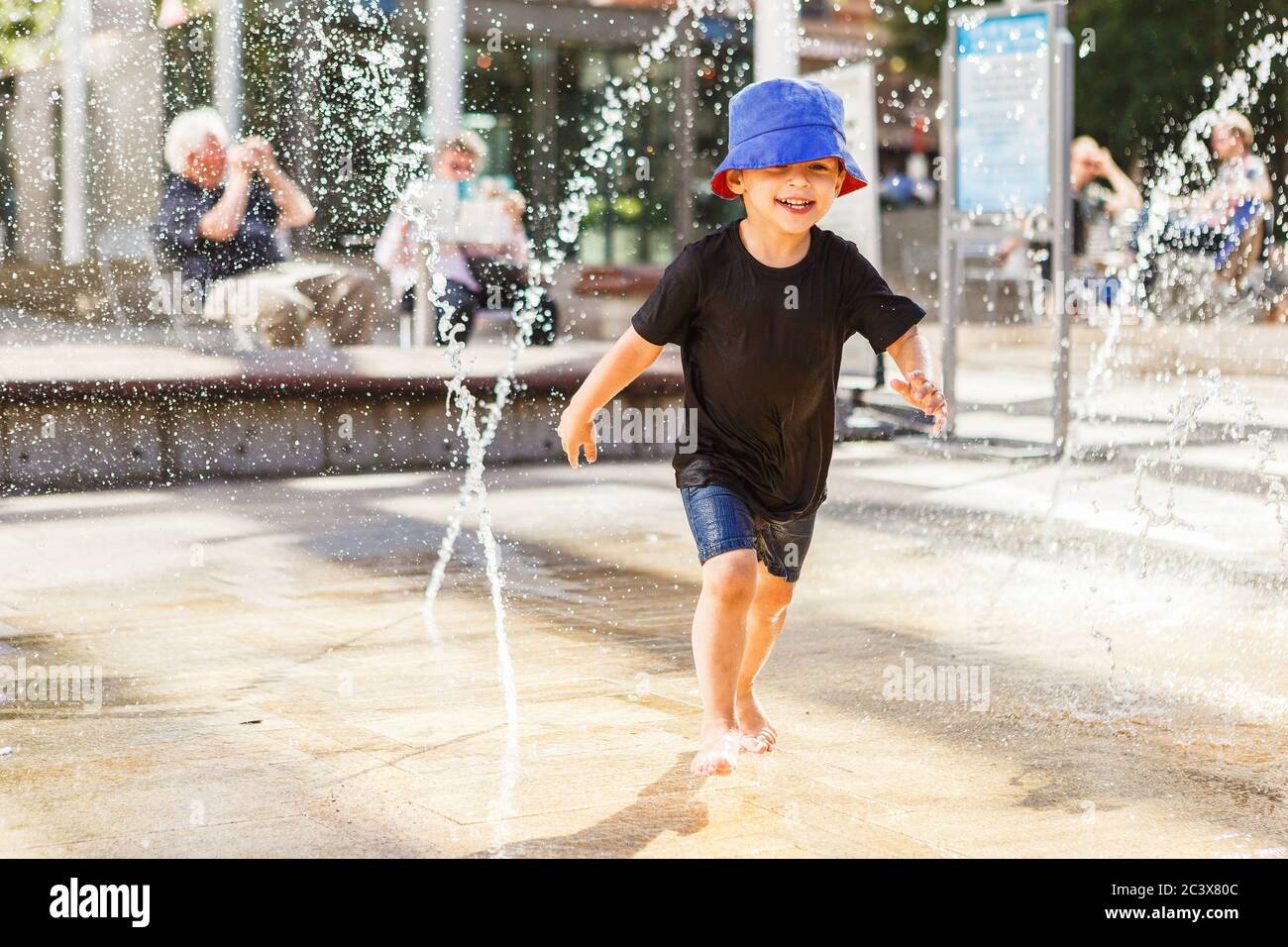 Happy little boy running between water jets in fountain on a heat ...