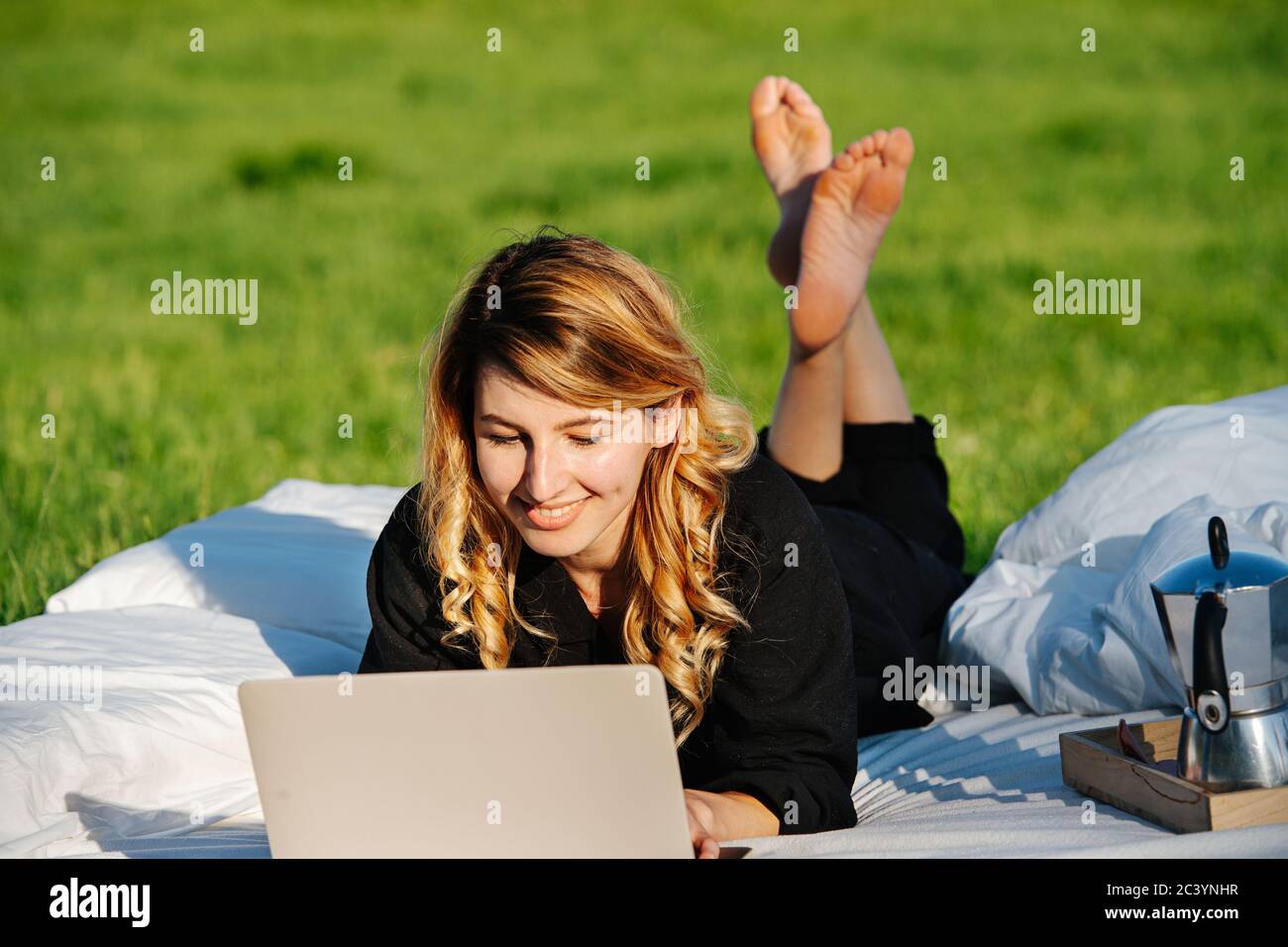 Charming woman working on a laptop, lying in bed outdoors, enjoying ...