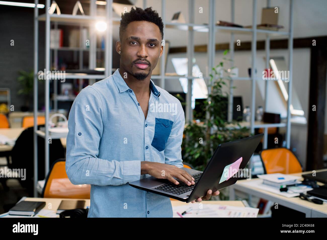 Confident African team leader, standing with laptop in modern creative ...