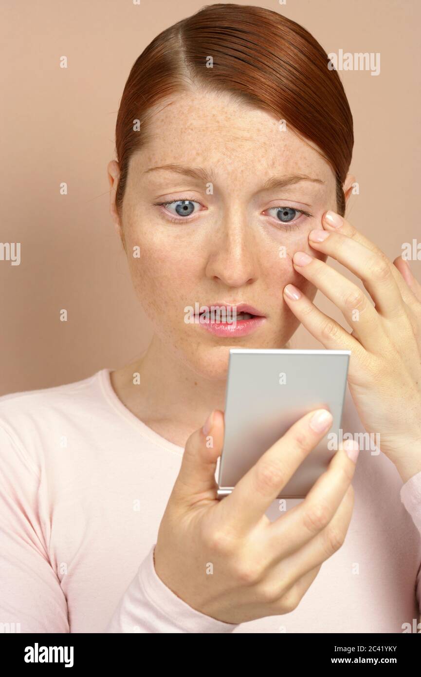 Young woman looks horrified at her face in a hand mirror - emotion ...