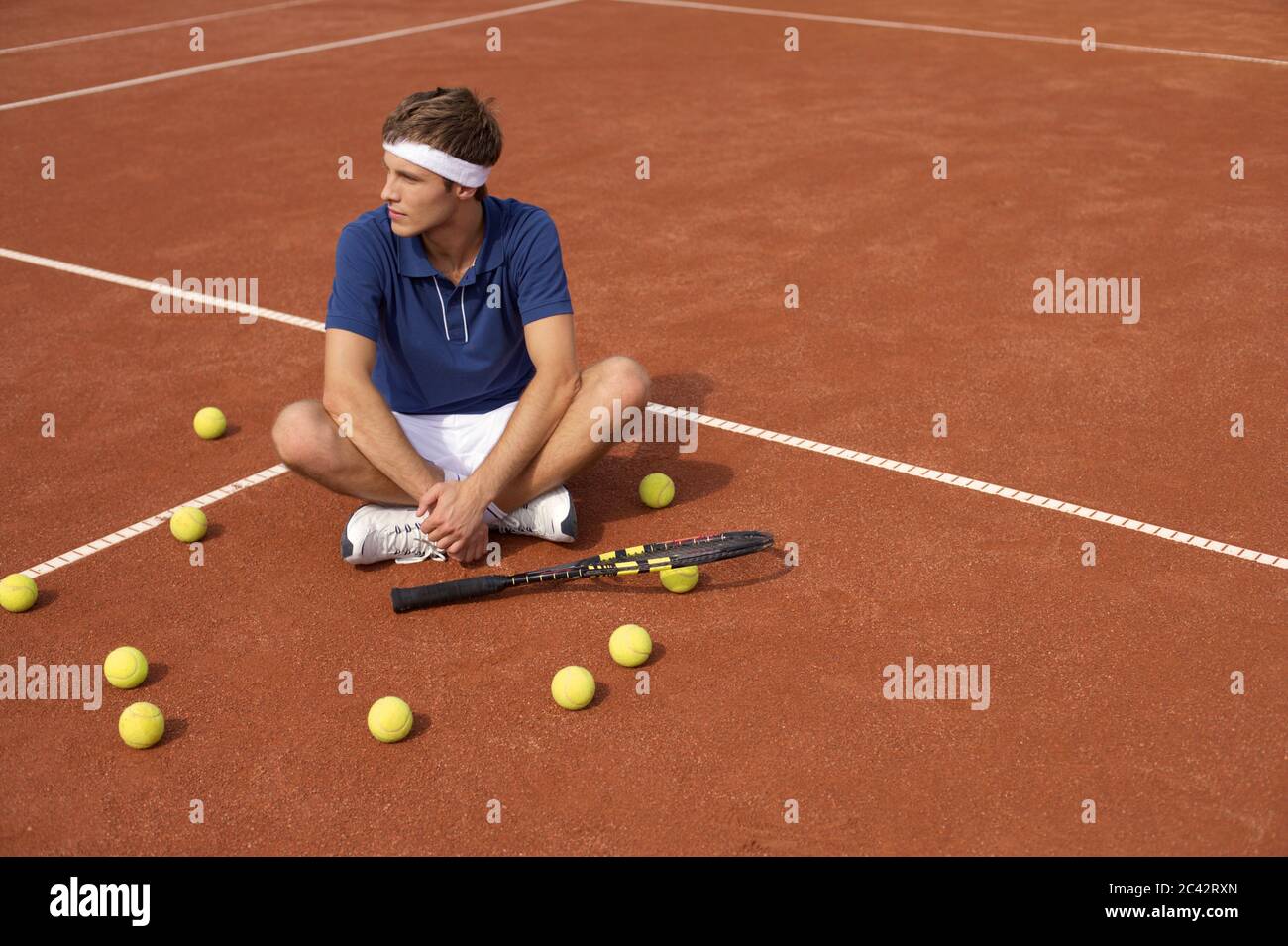 Young man sitting next to a tennis racket and balls on a sports field ...