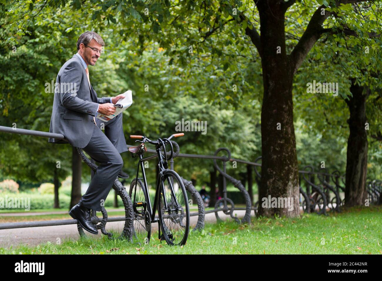 Businessman reading newspaper while sitting on railing in public park ...