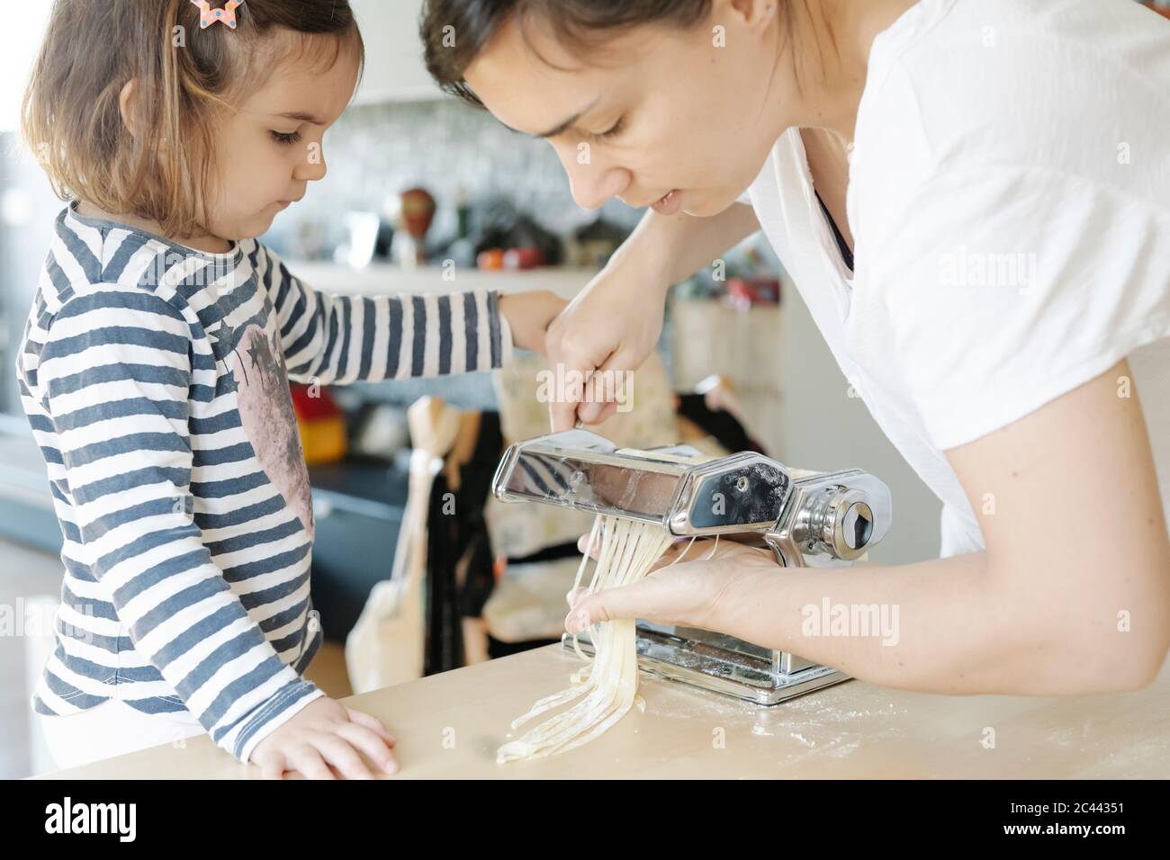 Cute girl looking at mother making spaghetti from equipment in kitchen ...