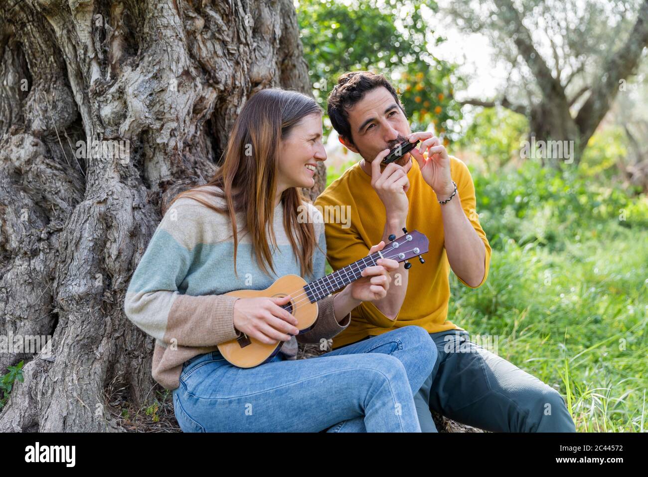 Happy couple playing musical instruments while sitting by tree trunk ...