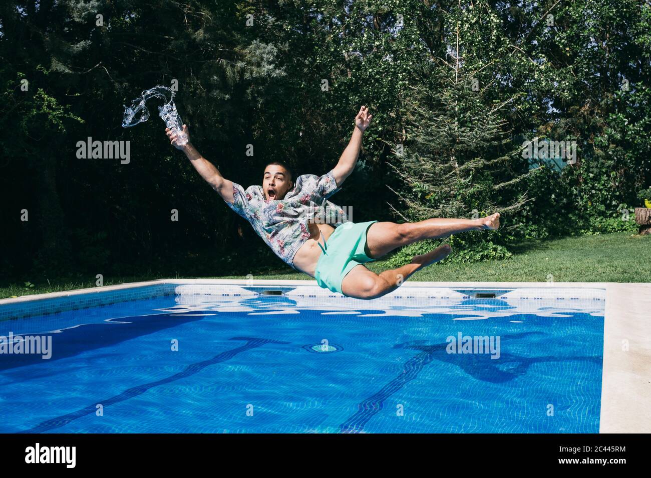 Shocked young man holding drink falling in swimming pool against trees ...