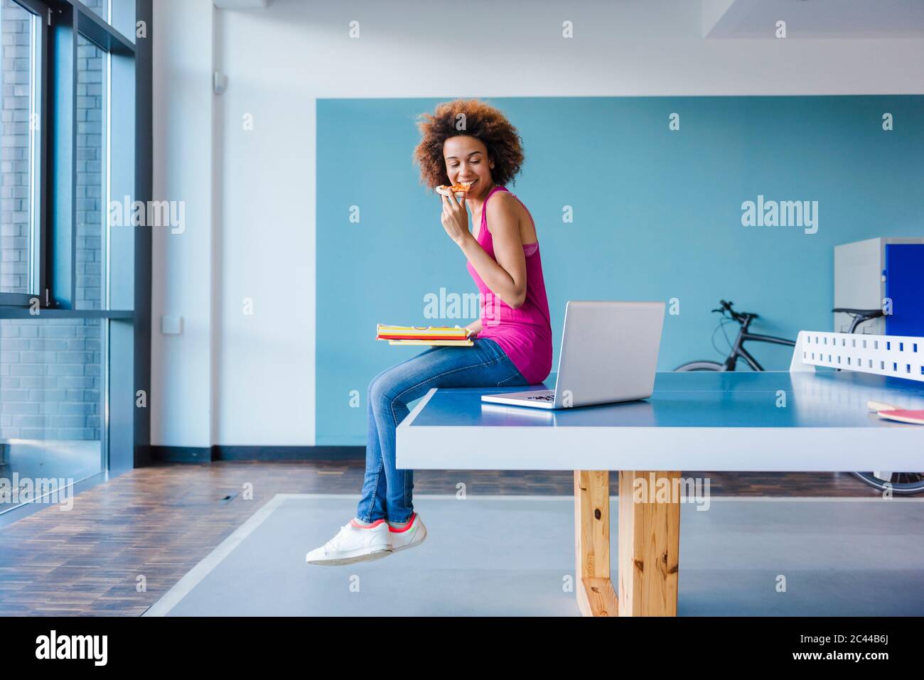 Young woman sitting on ping pong table, eating pizza, using laptop ...