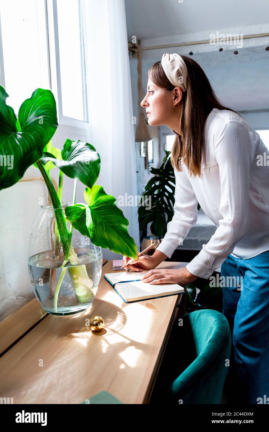 Woman looking through window while standing with diary at desk in ...