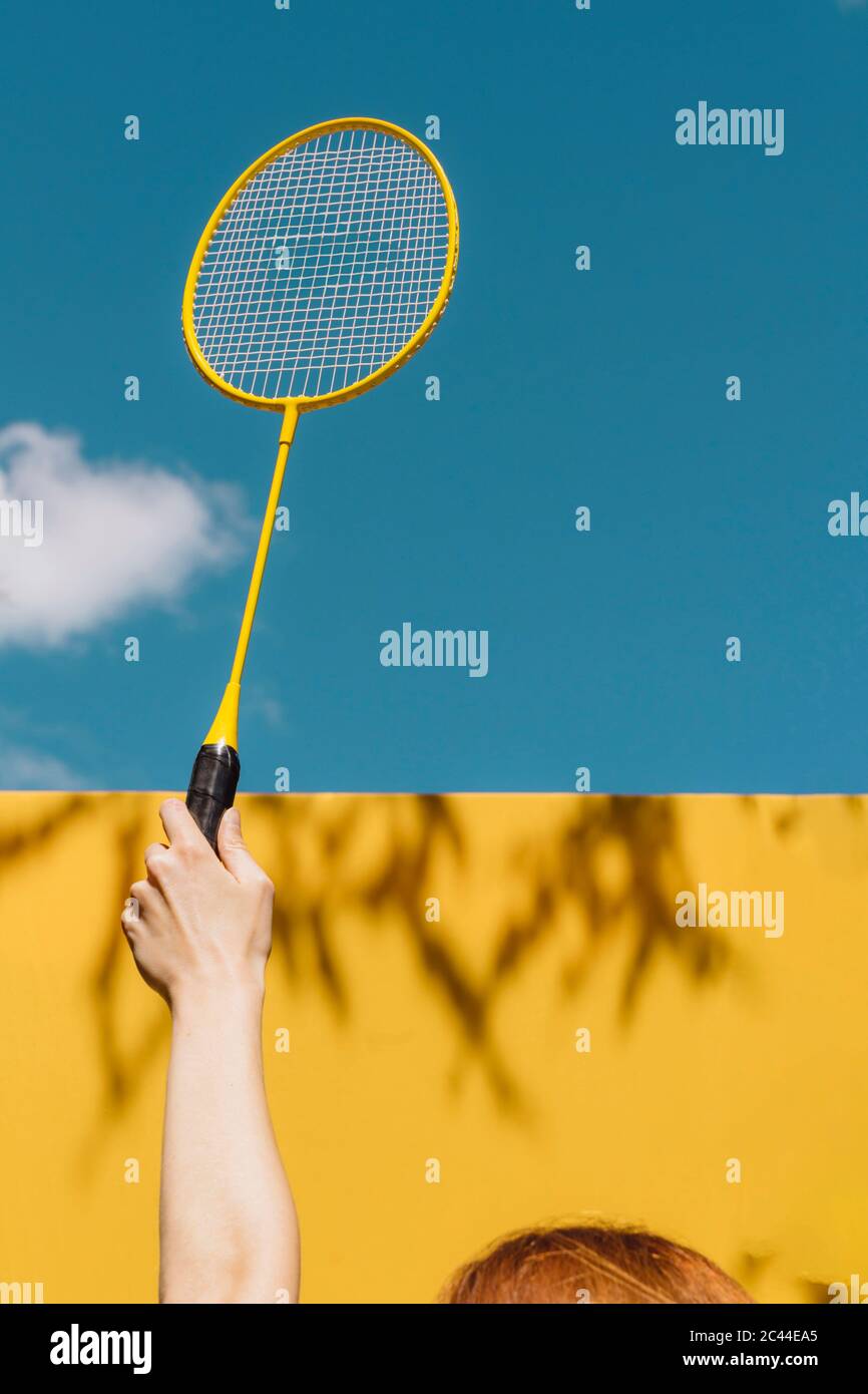 Woman's hand holding badminton racket over yellow wall against blue sky ...