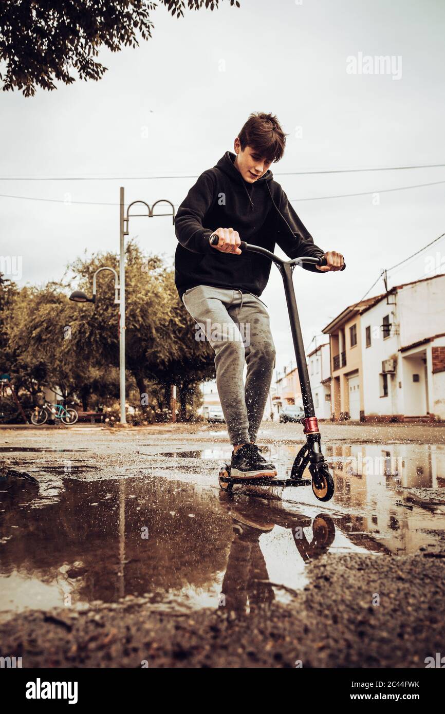 Teenage boy riding push scooter in puddle on street against sky Stock ...
