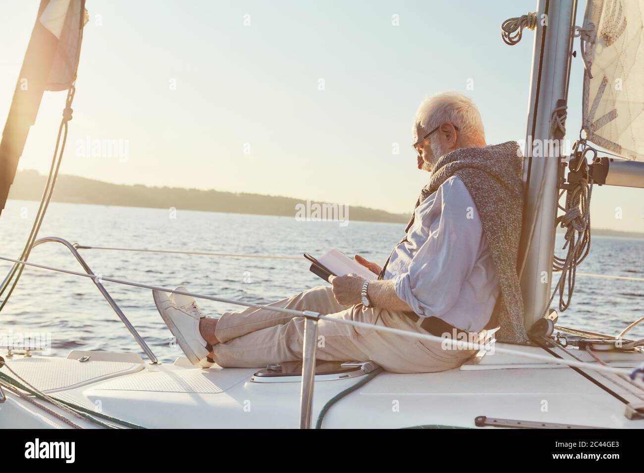 Relaxed senior man reading a book, while sitting on the side of sail ...