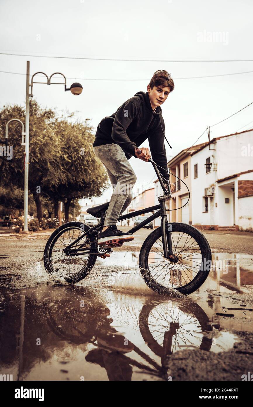 Confident teenage boy riding bicycle in puddle on street during rainy ...