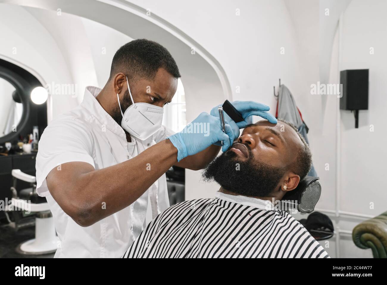 Man with full beard getting a shave, barber wearing surgical mask and ...