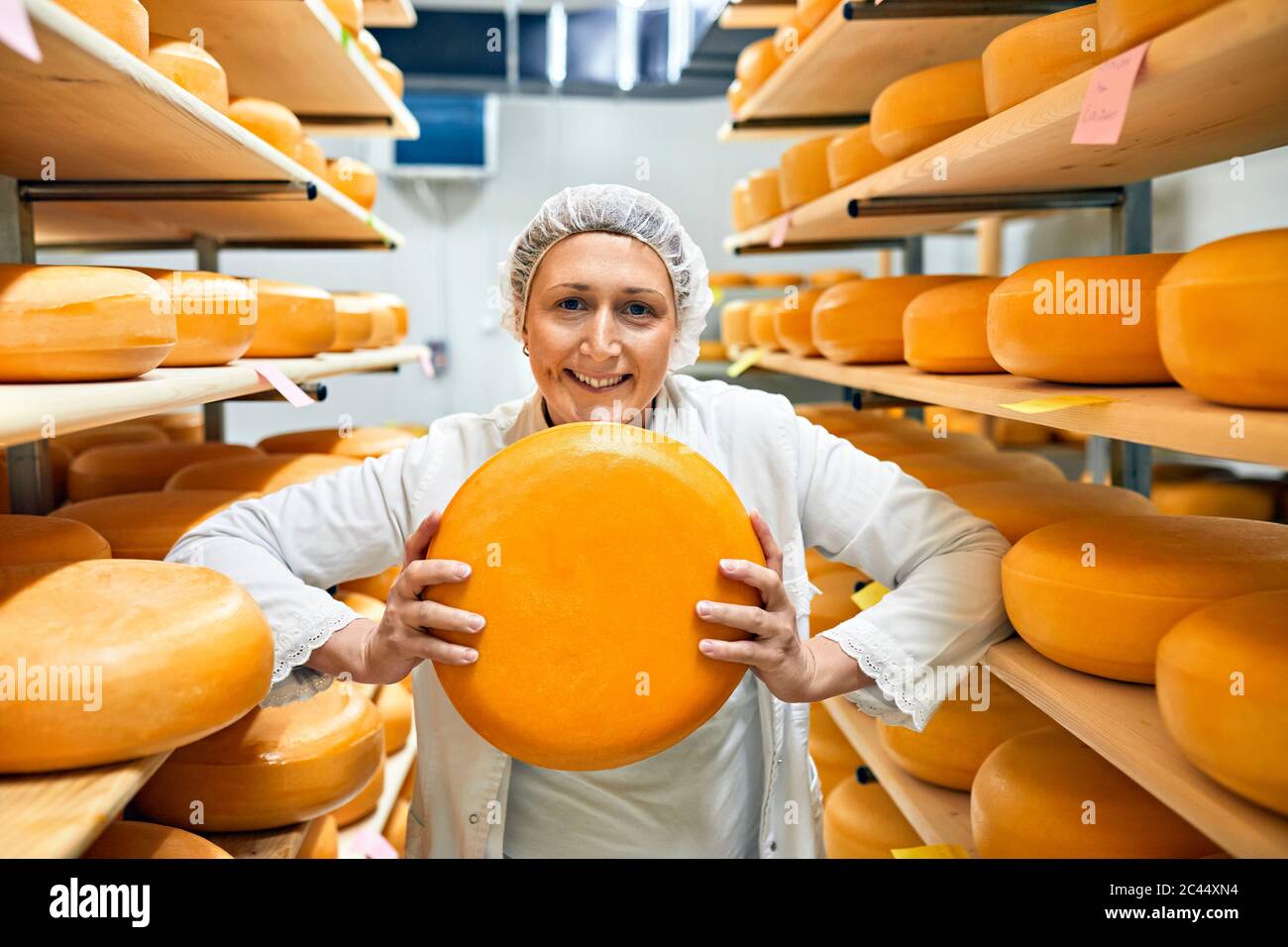 Cheese factory, smiling female worker with cheese wheel in storeroom ...