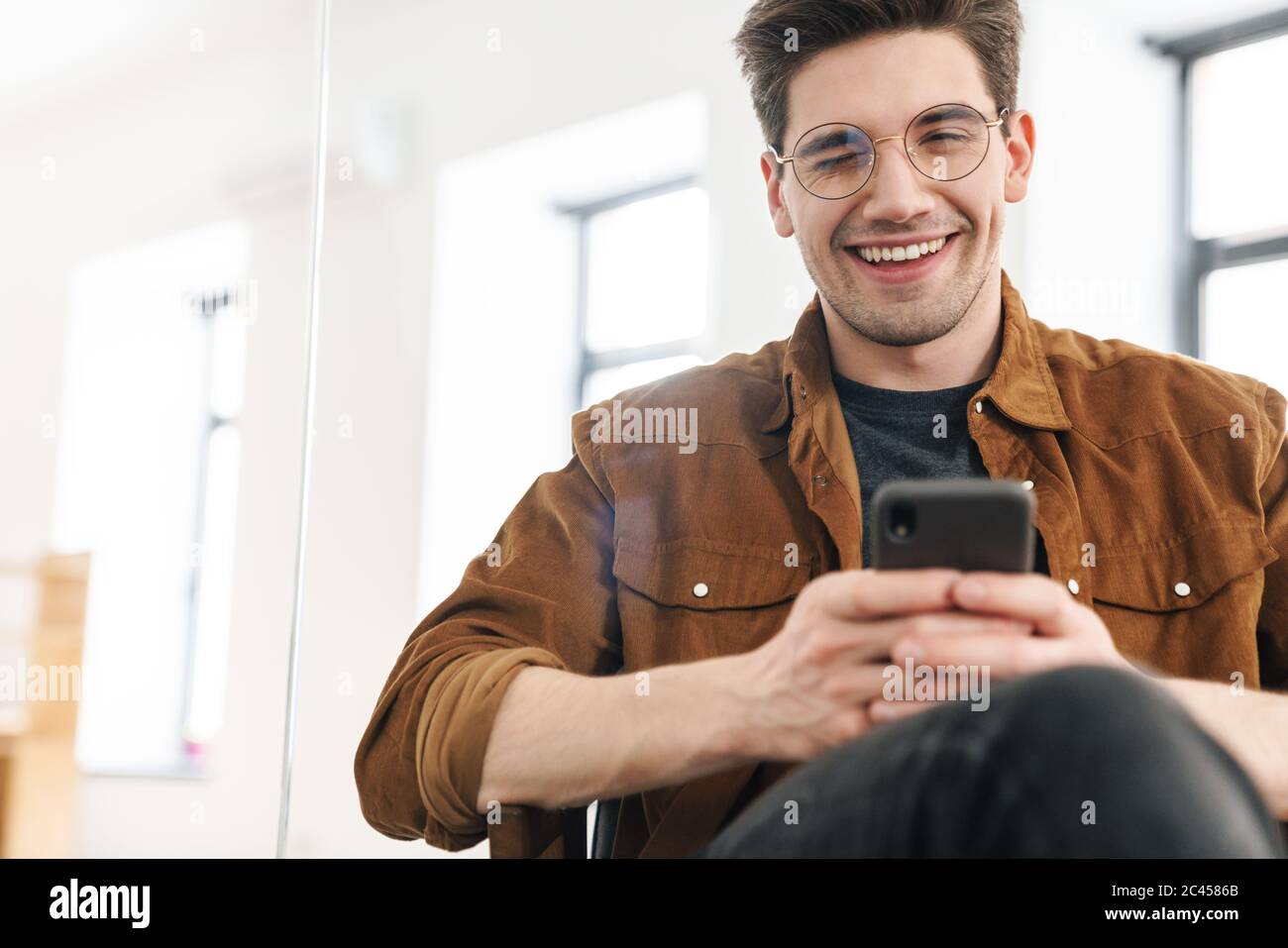 Image of handsome joyful young man winking and using cellphone while ...
