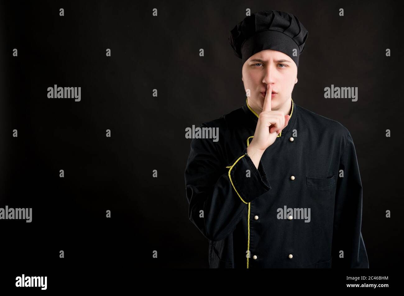 Portrait of young male dressed in a black chef suit showing shh gesture ...