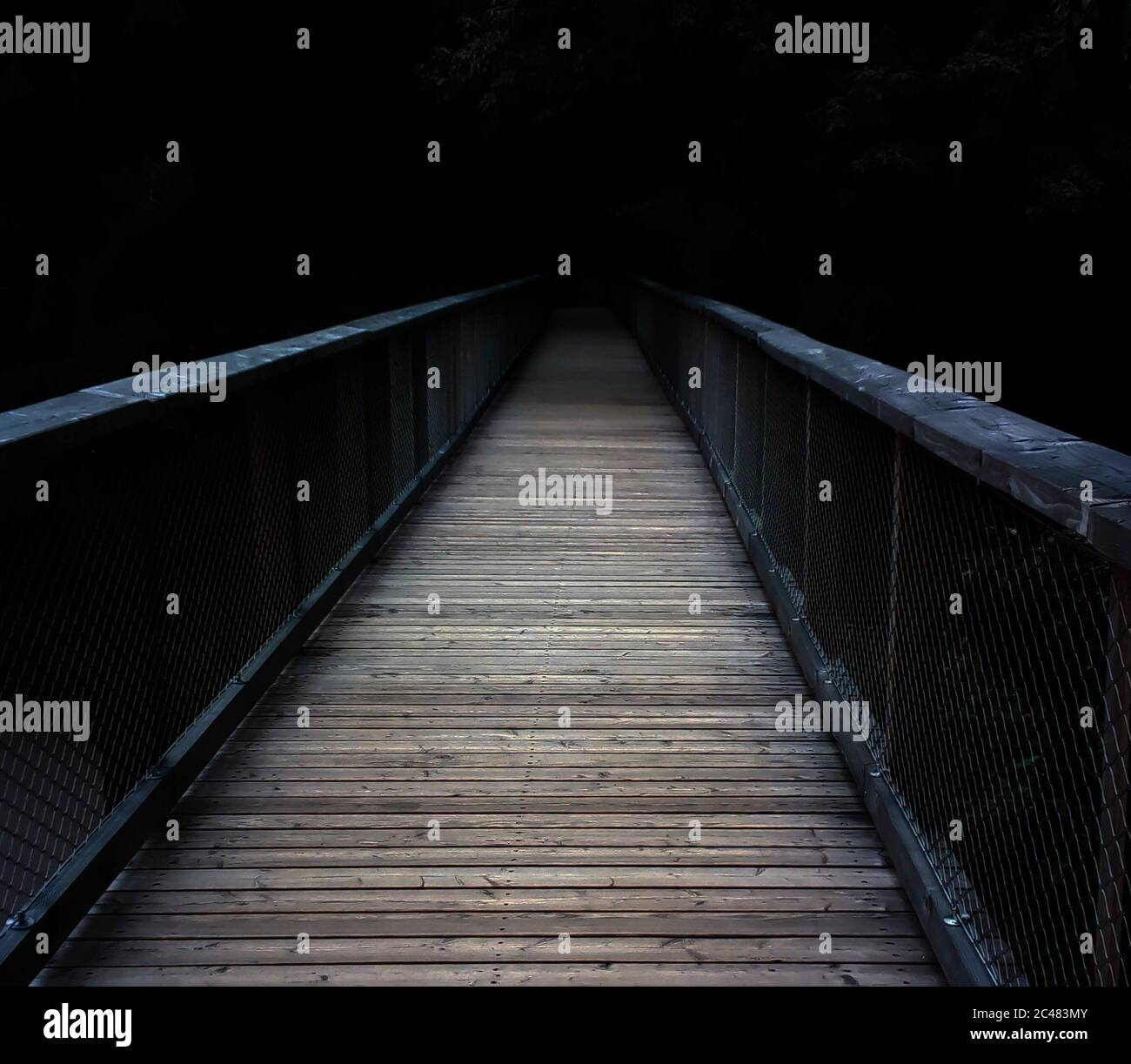 Wooden bridge with chain-link fences on the handrails leading to a dark ...