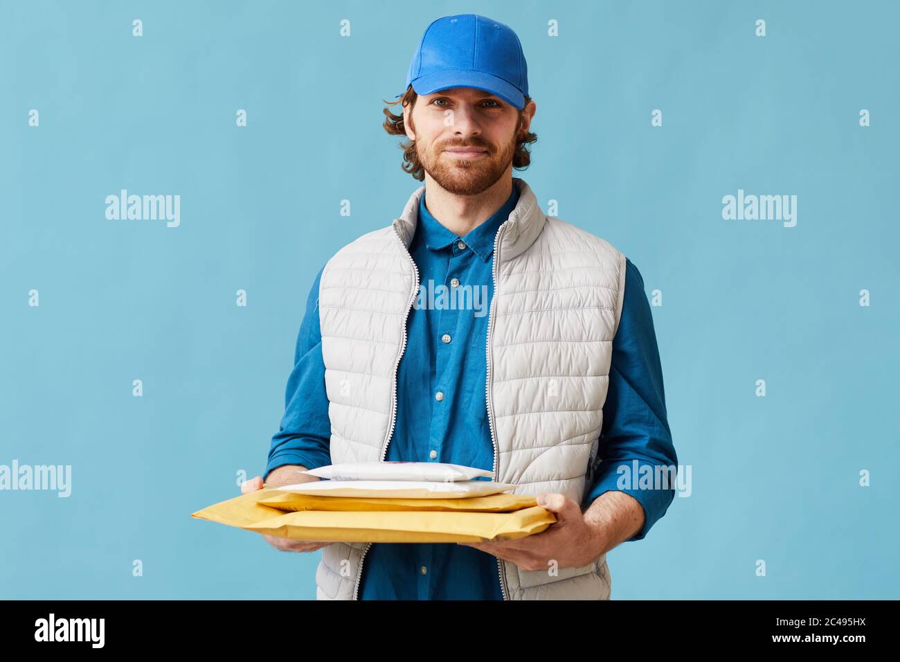 Portrait of young postman in uniform holding letters and looking at ...