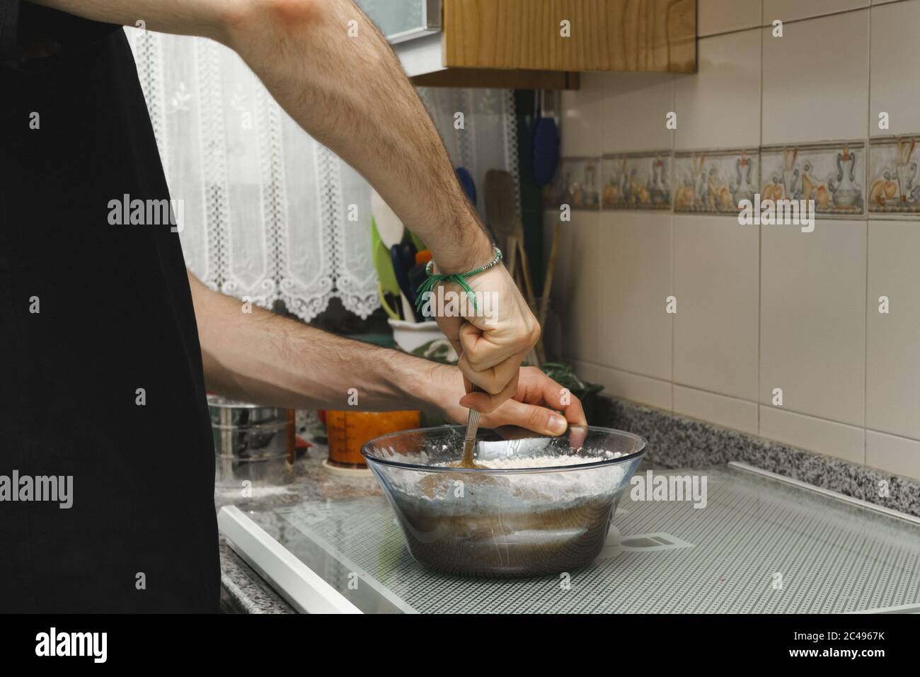 Man in the kitchen mixing the flour with a spoon in the big glass bowl ...