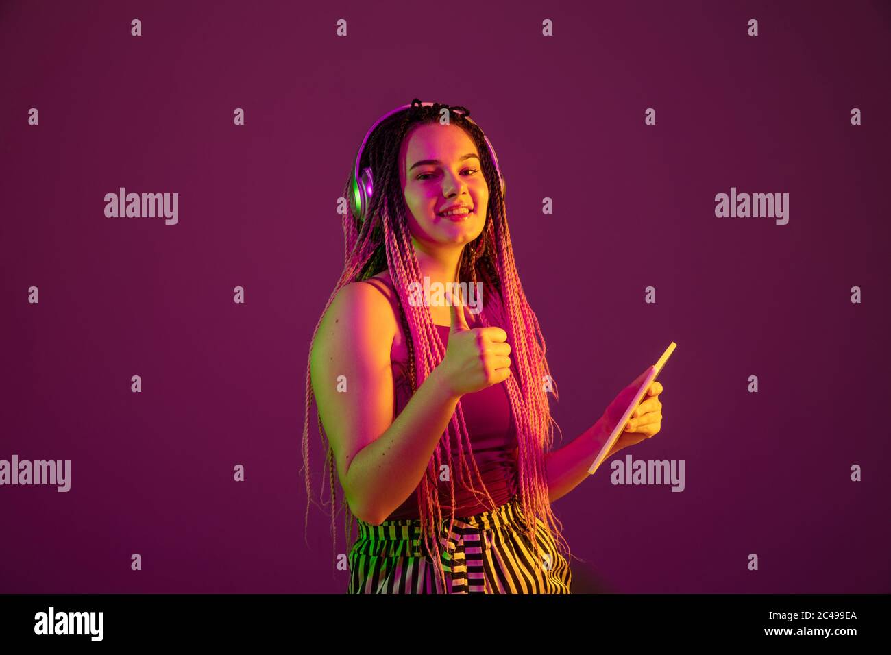 Thumb up. Young caucasian woman with headphones, tablet on pink studio ...