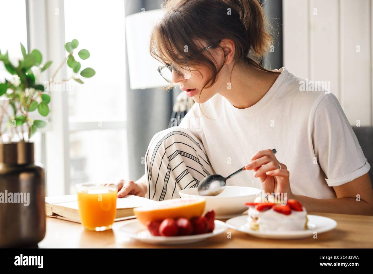 Photo of young focused woman reading book while having breakfast in ...