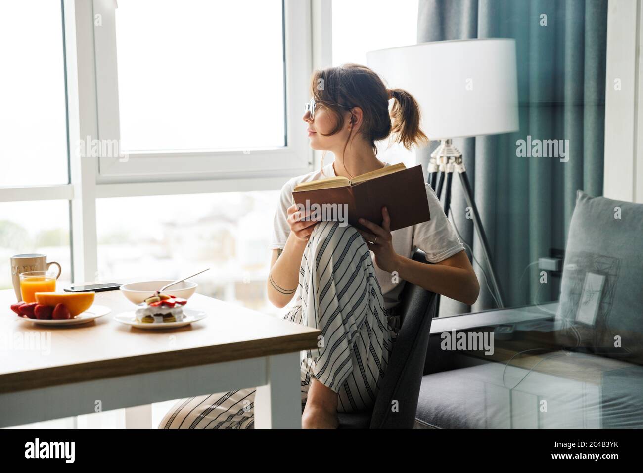 Photo of young concentrated woman reading book while having breakfast ...