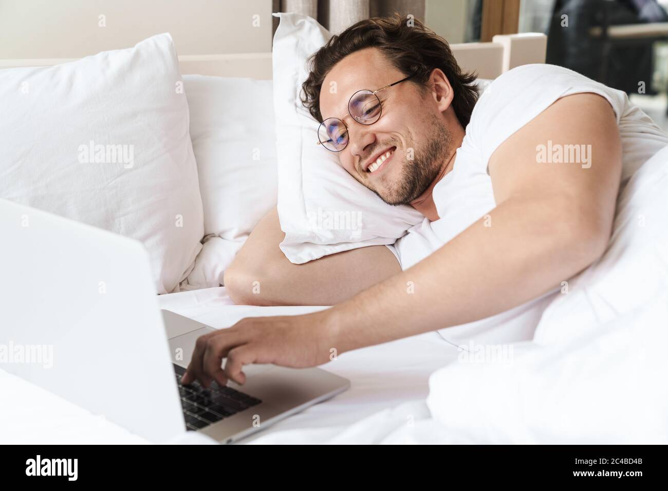 Smiling young man laying on a pillow in bed, using laptop computer ...