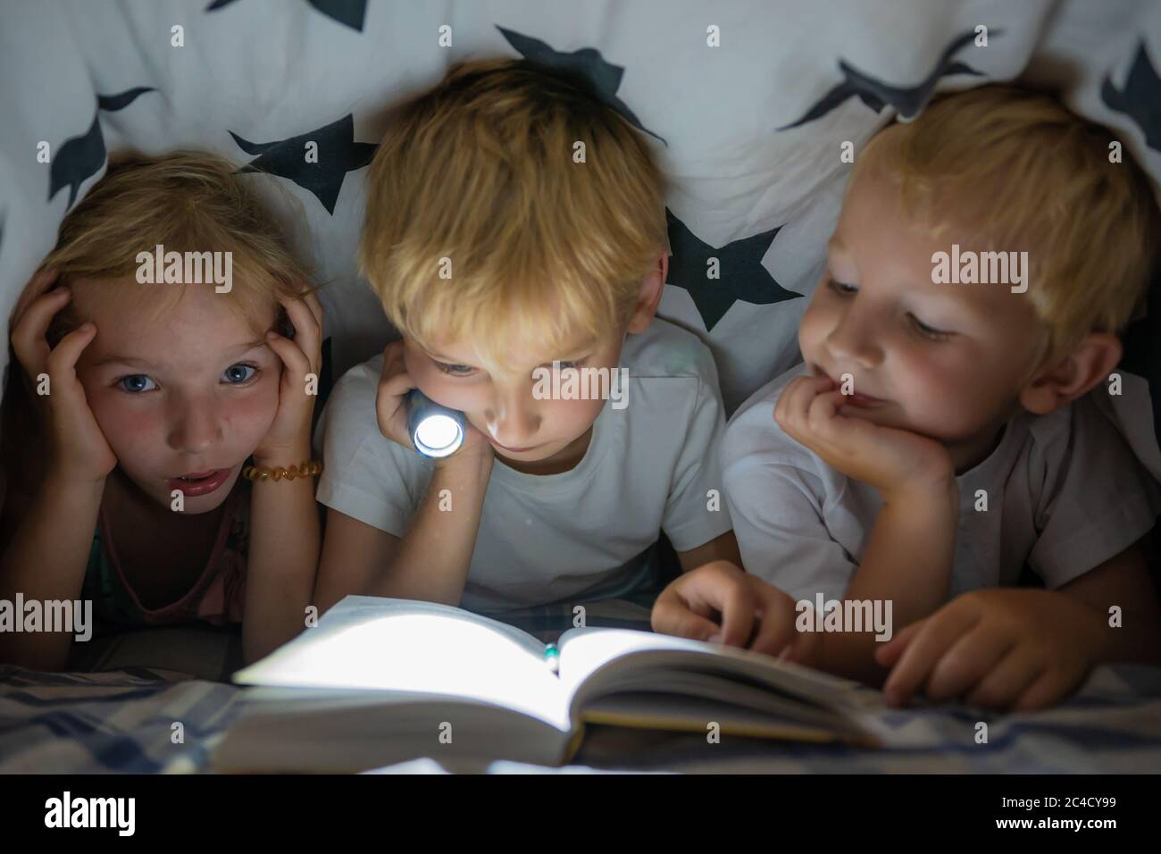 Three young children are reading a book with a flashlight under the ...