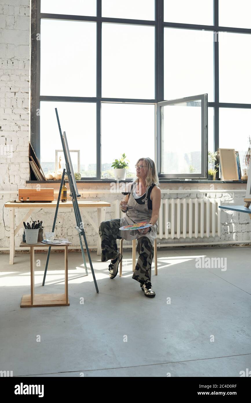 Relaxed female artist in apron holding palette and drinking wine while ...