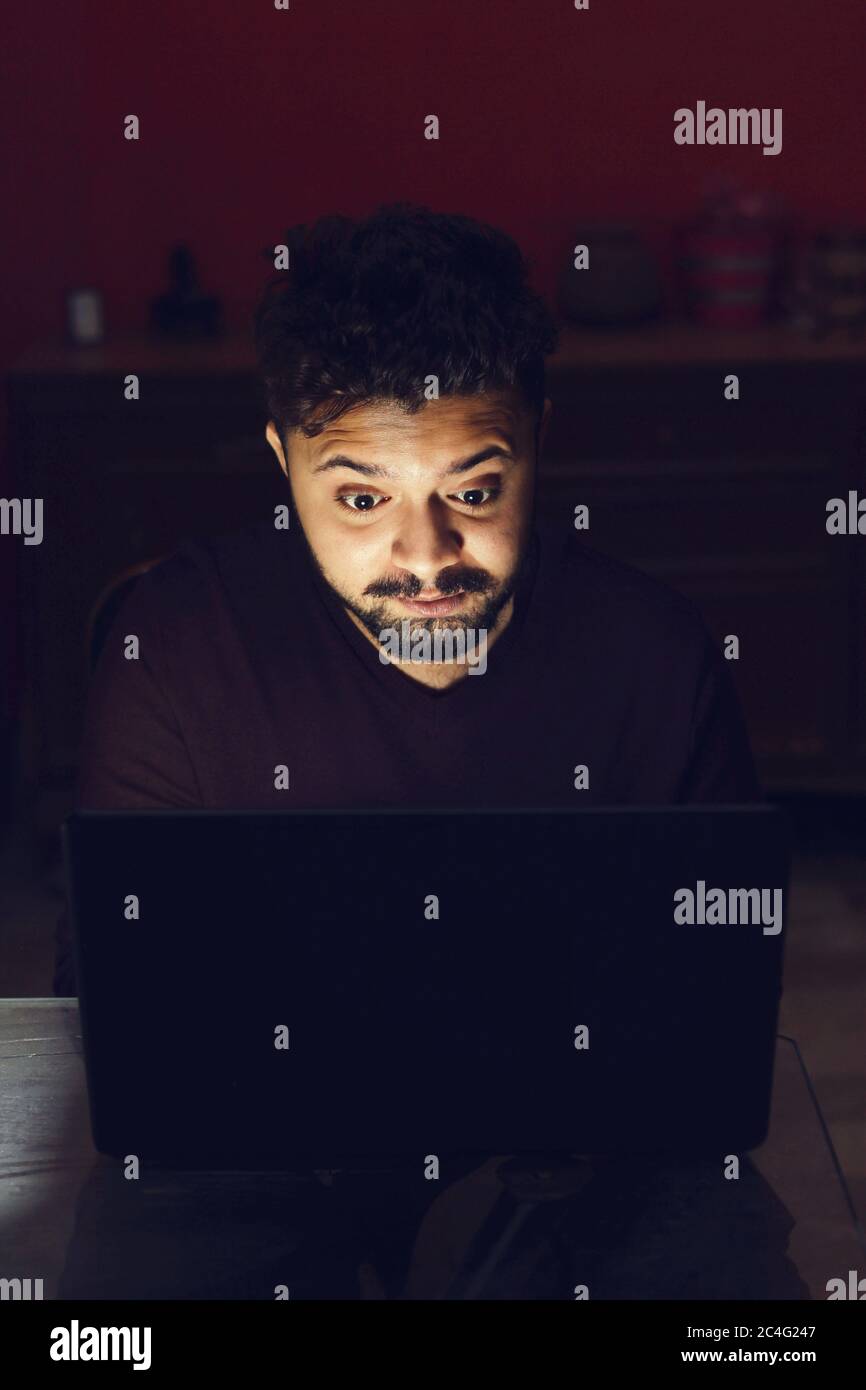 Young man looking at laptop computer in dark room, browsing on internet ...