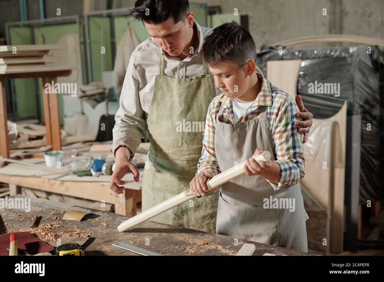 Teenage boy in apron making plank smooth with sandpaper under control ...