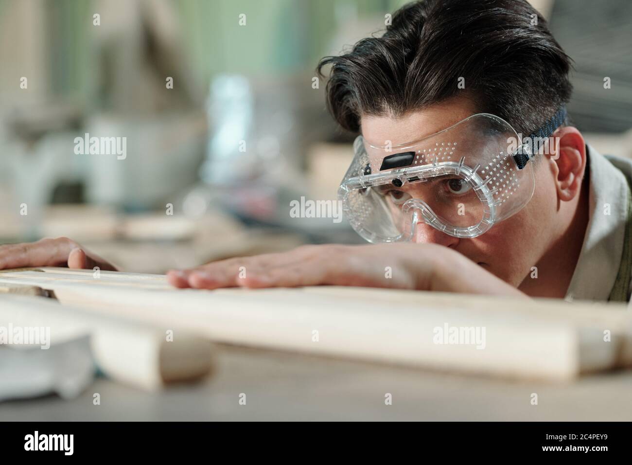Focused brunette male carpenter in safety goggles checking processed ...