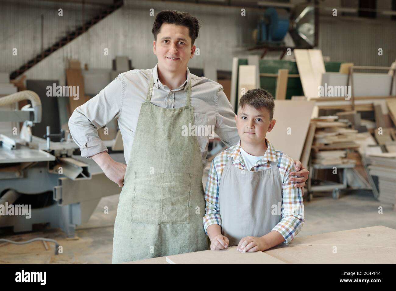 Portrait of smiling middle-aged carpenter in apron embracing teenage ...